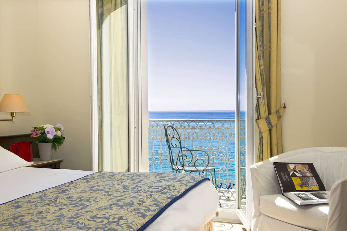 Quarto de hotel com uma cama, uma cadeira junto à janela e vista para o mar.