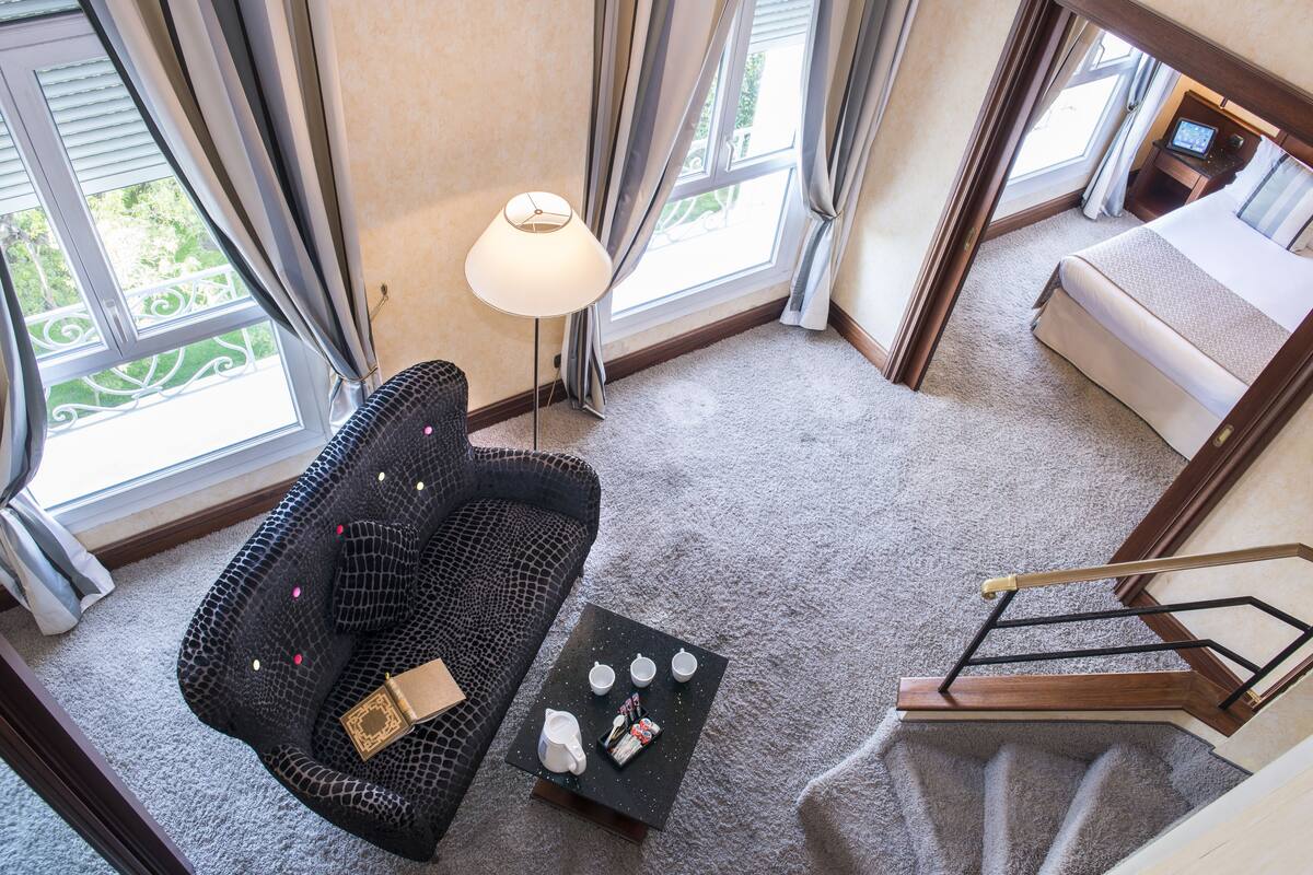 Quarto de hotel com sofá estampado em preto, uma pequena mesa de centro com xícaras e um livro, um abajur e vista para o exterior através de grandes janelas.