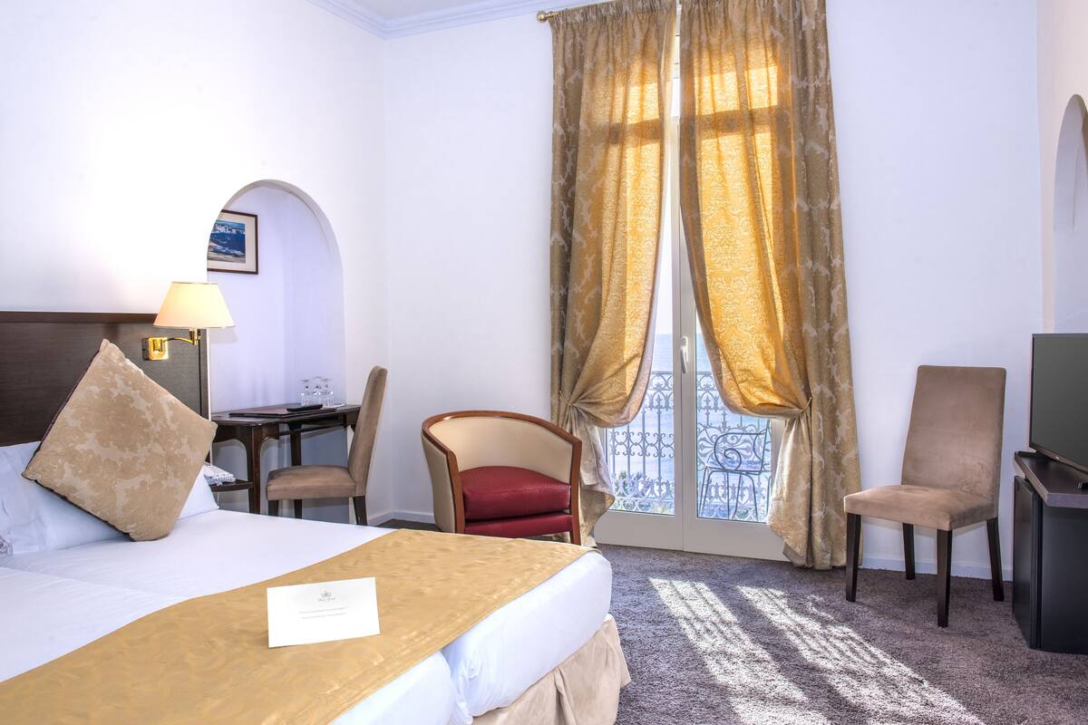 Quarto de hotel com cama, duas cadeiras, escrivaninha e televisão.