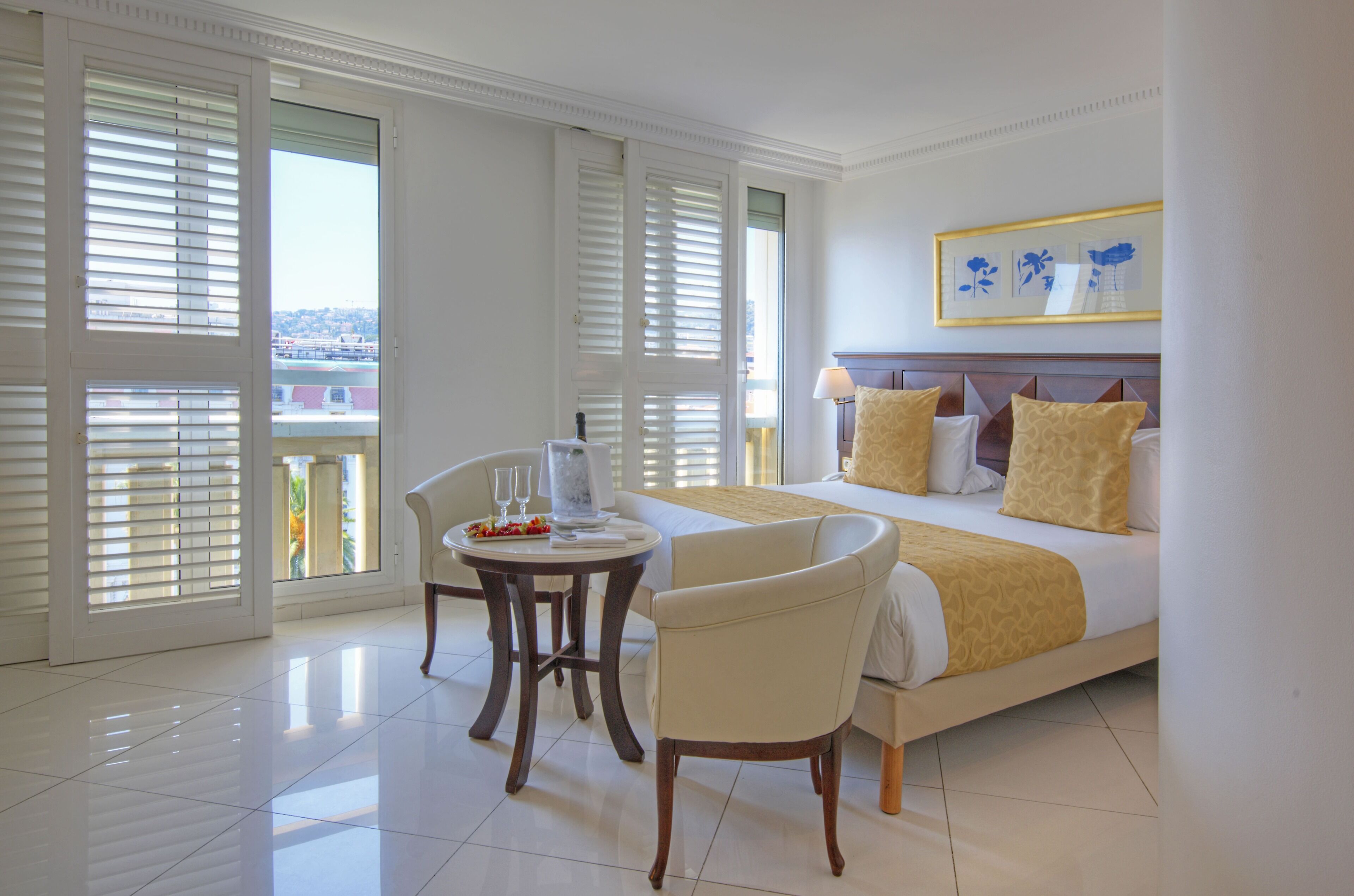 Quarto de hotel com uma cama, duas cadeiras, uma mesinha com copos e um jarro, e uma janela com persianas.