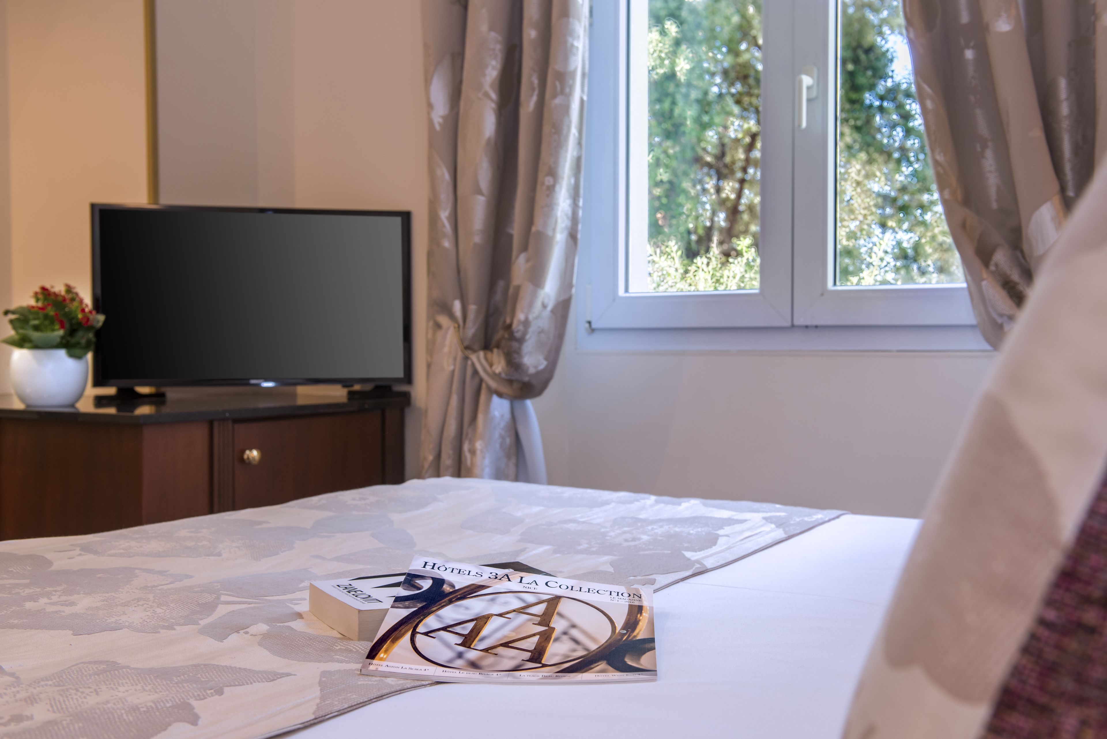 Quarto de hotel com cama, televisão sobre um móvel de madeira, janela com cortinas e um livro intitulado 
