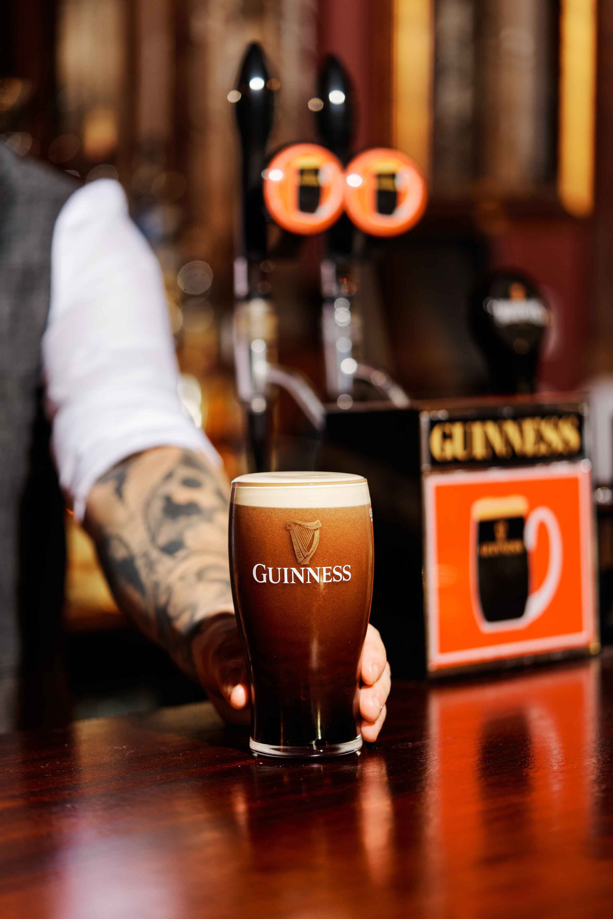 Um copo de cerveja Guinness com logotipo, segurado por uma pessoa com tatuagens, em frente a uma torneira e uma caixa com a inscrição "GUINNESS".
