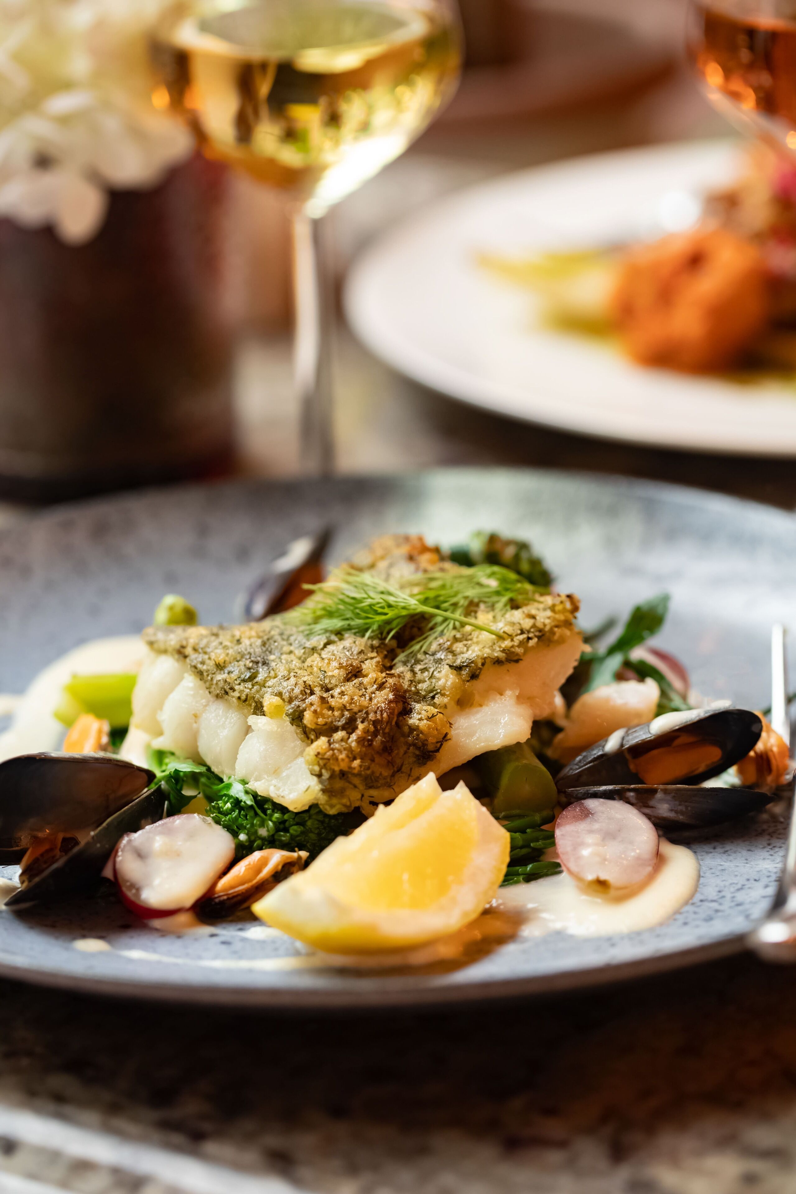 Um prato de frutos do mar com mexilhões, camarões e um pedaço de peixe, acompanhado de uma rodela de limão e um copo de vinho branco.