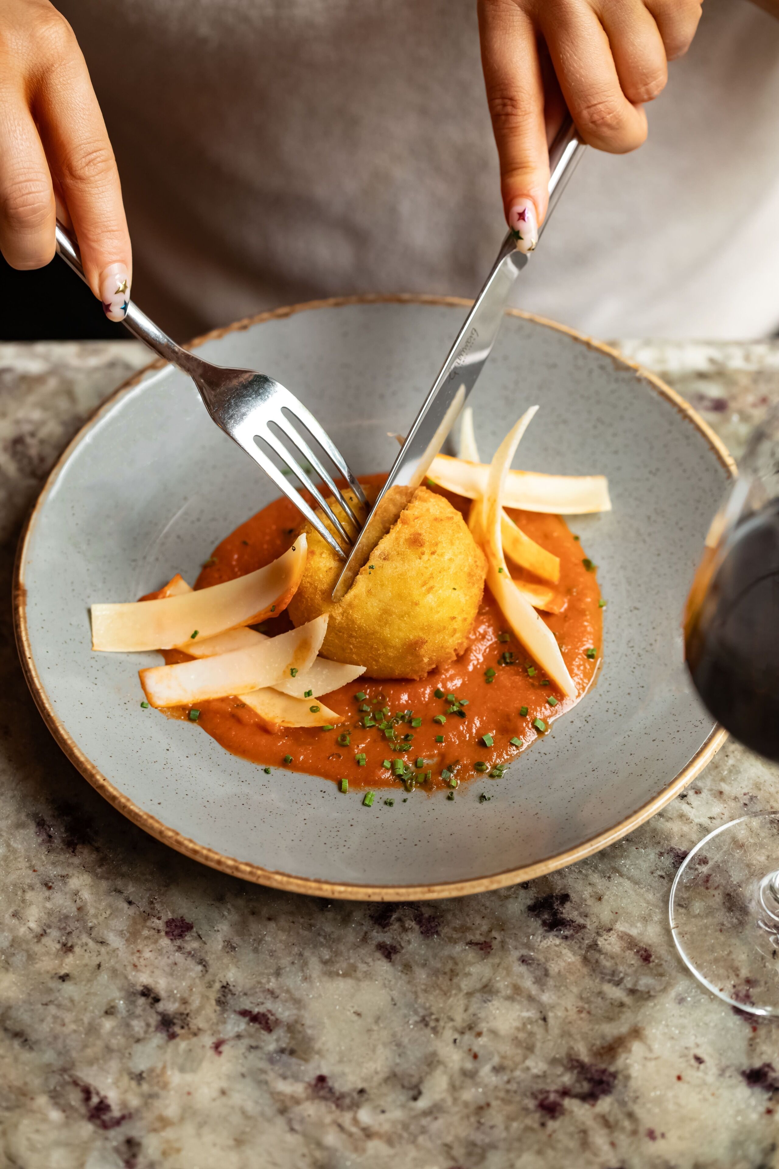Uma pessoa usando garfo e faca para cortar um prato com massa e um item dourado, possivelmente um croquete, em um prato salpicado.