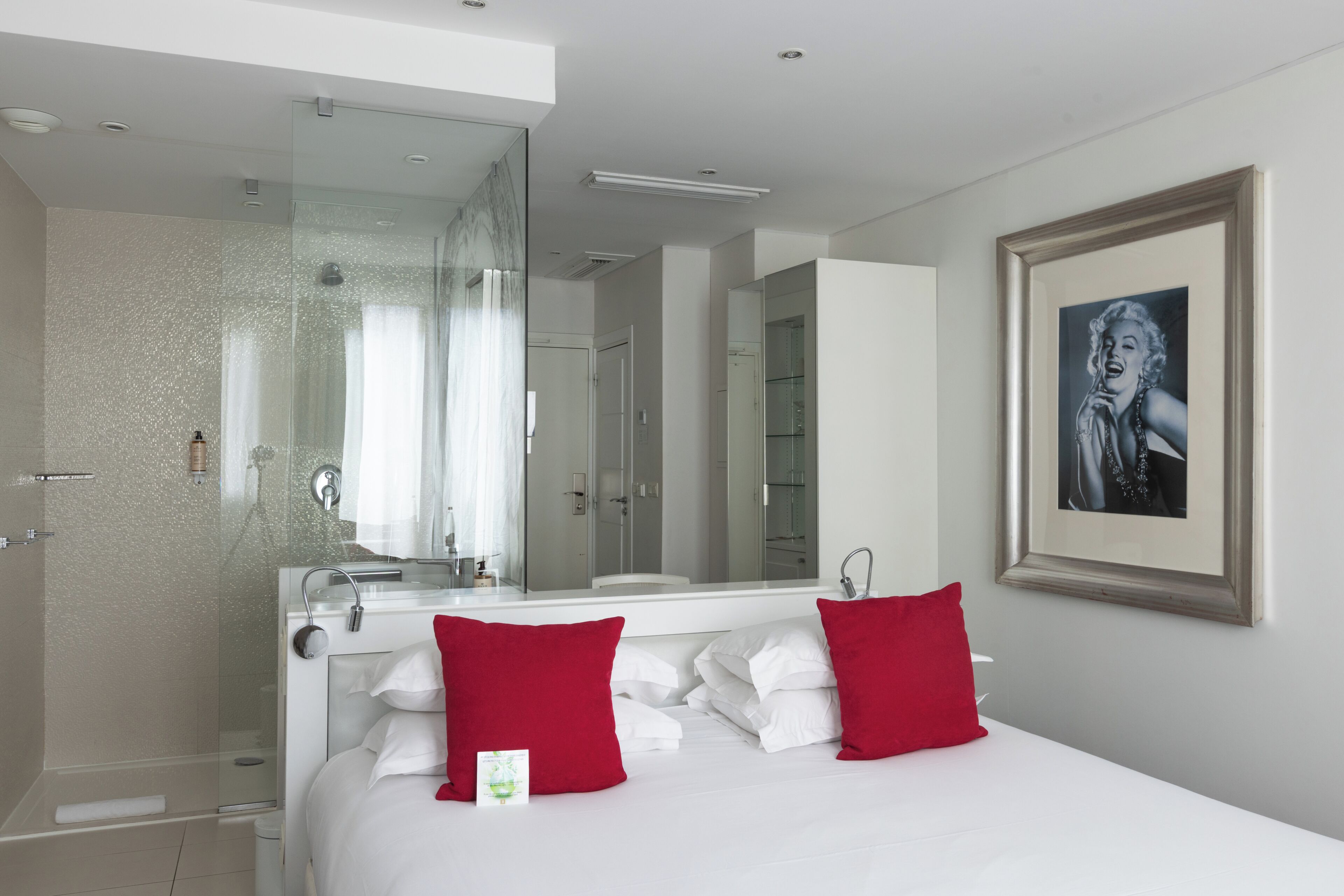 Quarto de hotel moderno com uma cama grande, área de chuveiro, banheiro com box de vidro e uma moldura com imagem na parede.