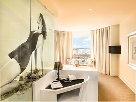 Quarto de hotel com uma janela grande, uma mesa com telefone, uma lâmpada e uma cama com vista para a paisagem urbana.