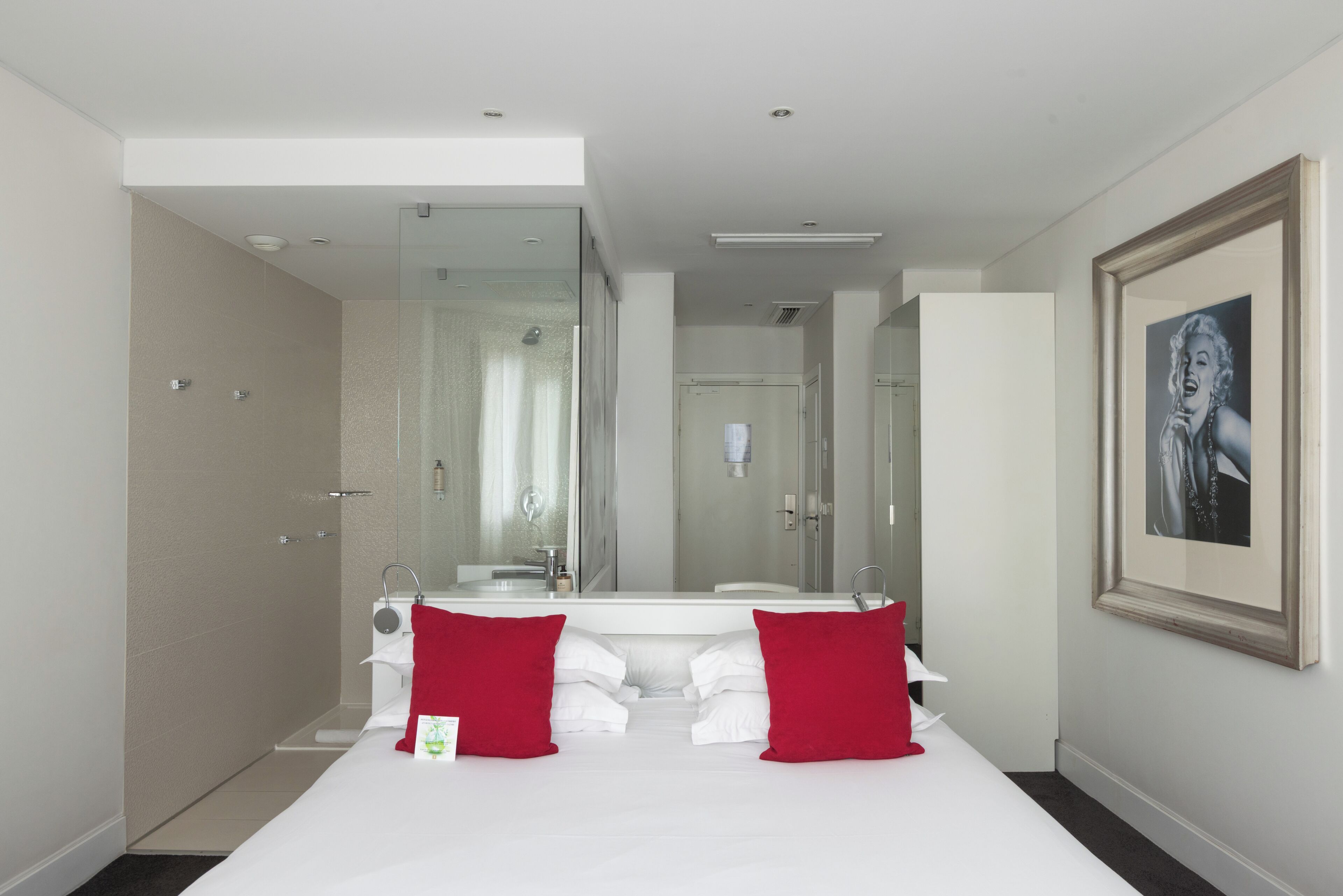 Quarto de hotel moderno com uma cama grande, box de vidro para o chuveiro, um espelho amplo e um quadro decorativo na parede.