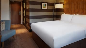 Quarto de hotel com uma cama grande, uma poltrona azul, um painel de cabeceira de madeira e um quadro pendurado na parede.