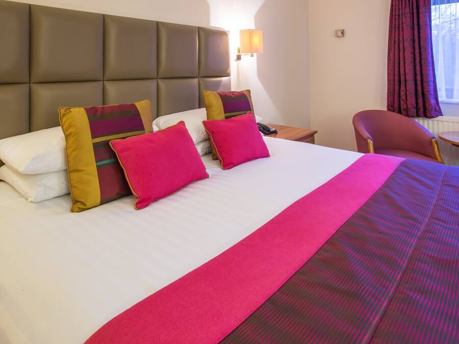 Quarto de hotel com uma cama grande, várias almofadas, mesa de cabeceira, uma cadeira e janela com cortinas.