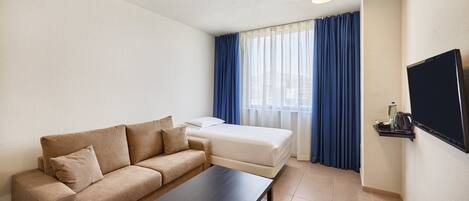 Quarto de hotel moderno com sofá, mesa pequena, televisão e janela com cortinas.