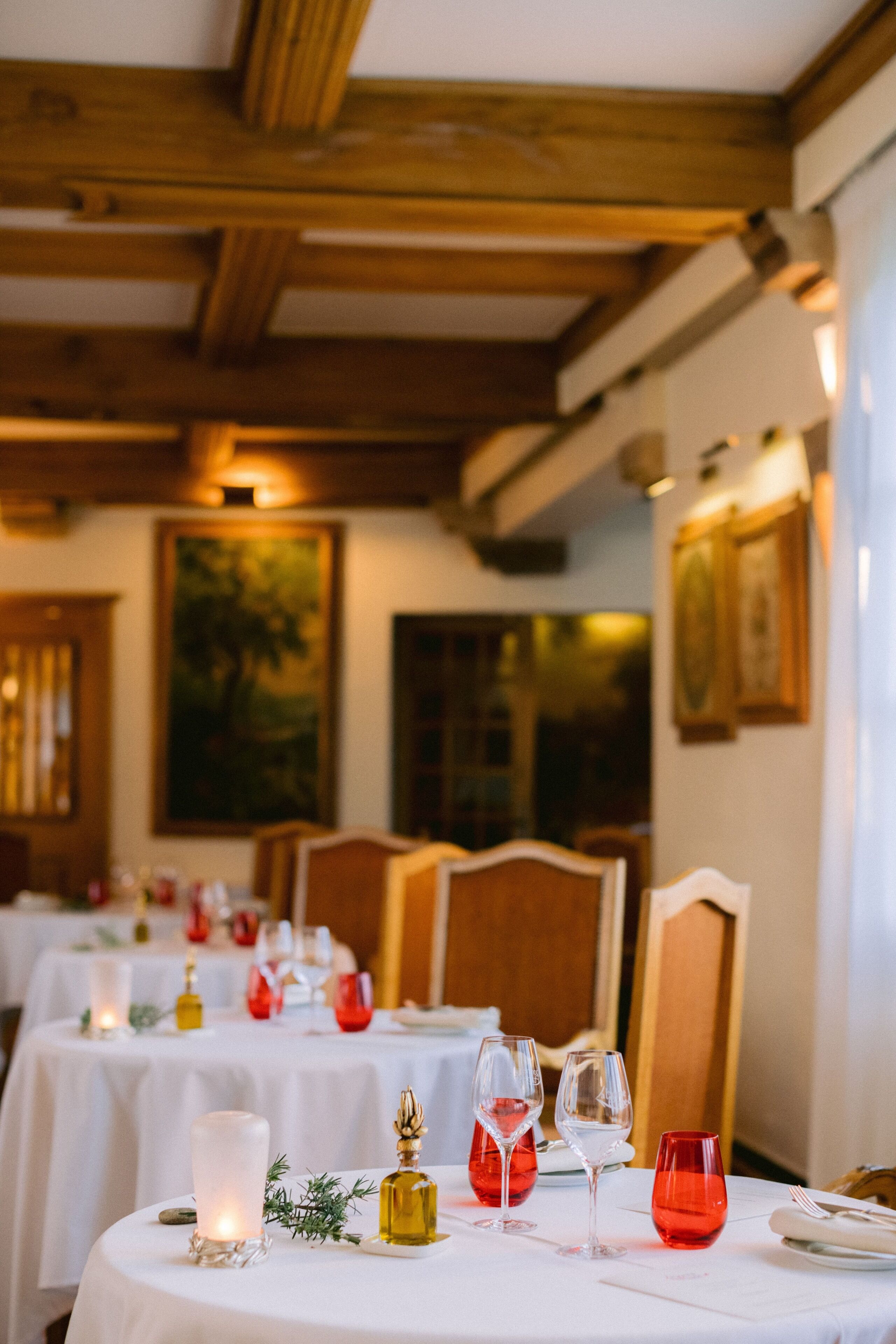 Uma sala de jantar com mesas redondas preparadas para refeição, com toalhas brancas, copos vermelhos e velas.