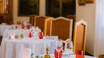 Uma sala de jantar com mesas redondas preparadas para refeição, com toalhas brancas, copos vermelhos e velas.