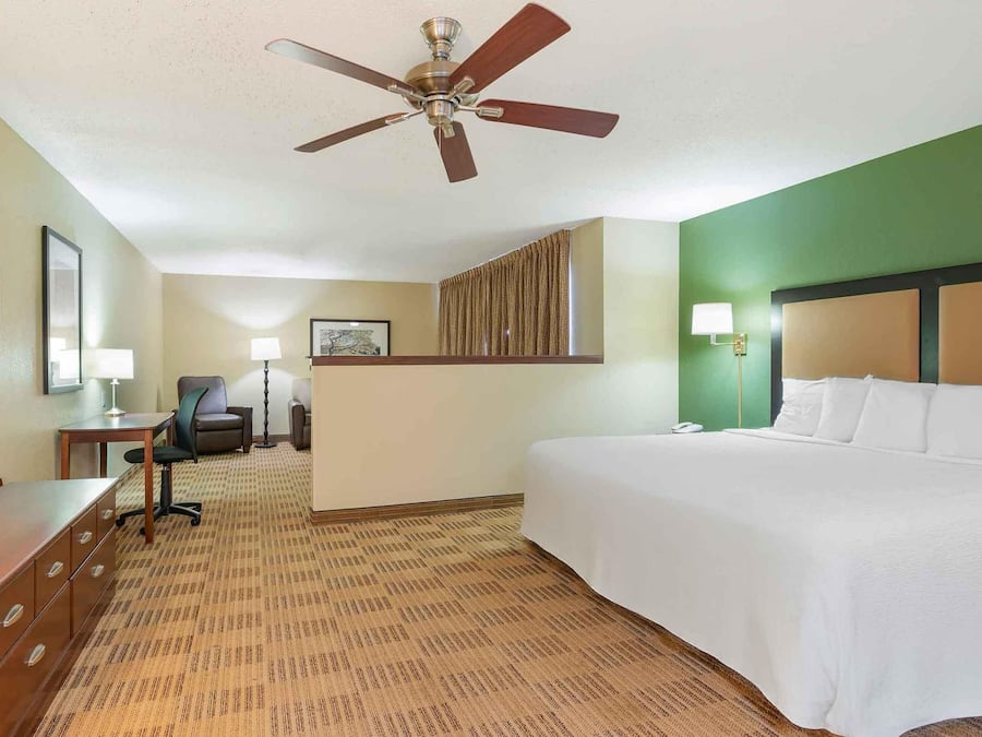 Quarto de hotel com cama, escrivaninha, cadeira, ventilador de teto e televisão na parede.