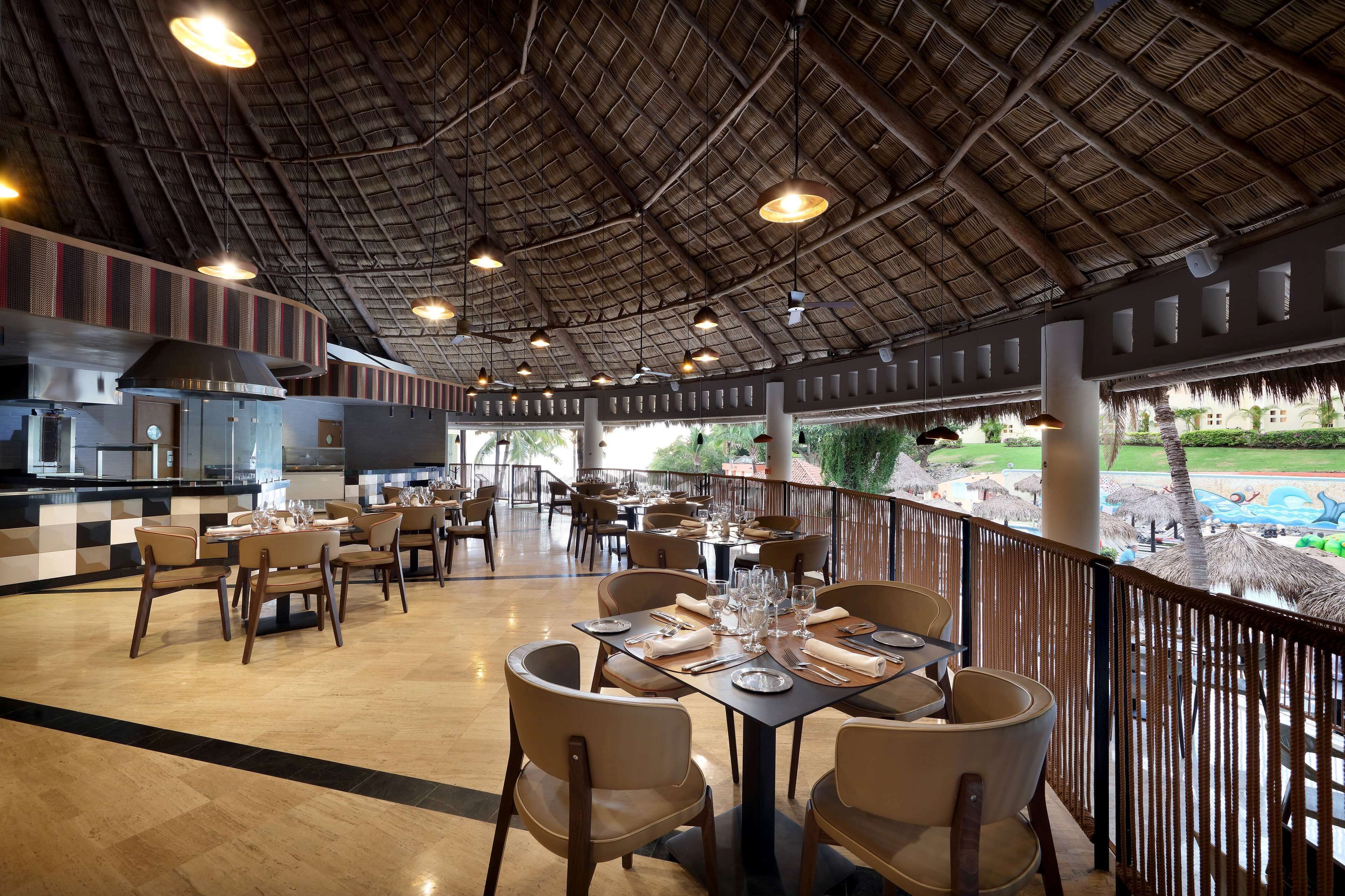 Um restaurante espaçoso com um telhado alto de palha, várias mesas de jantar com cadeiras e vista para uma área de piscina ao ar livre.