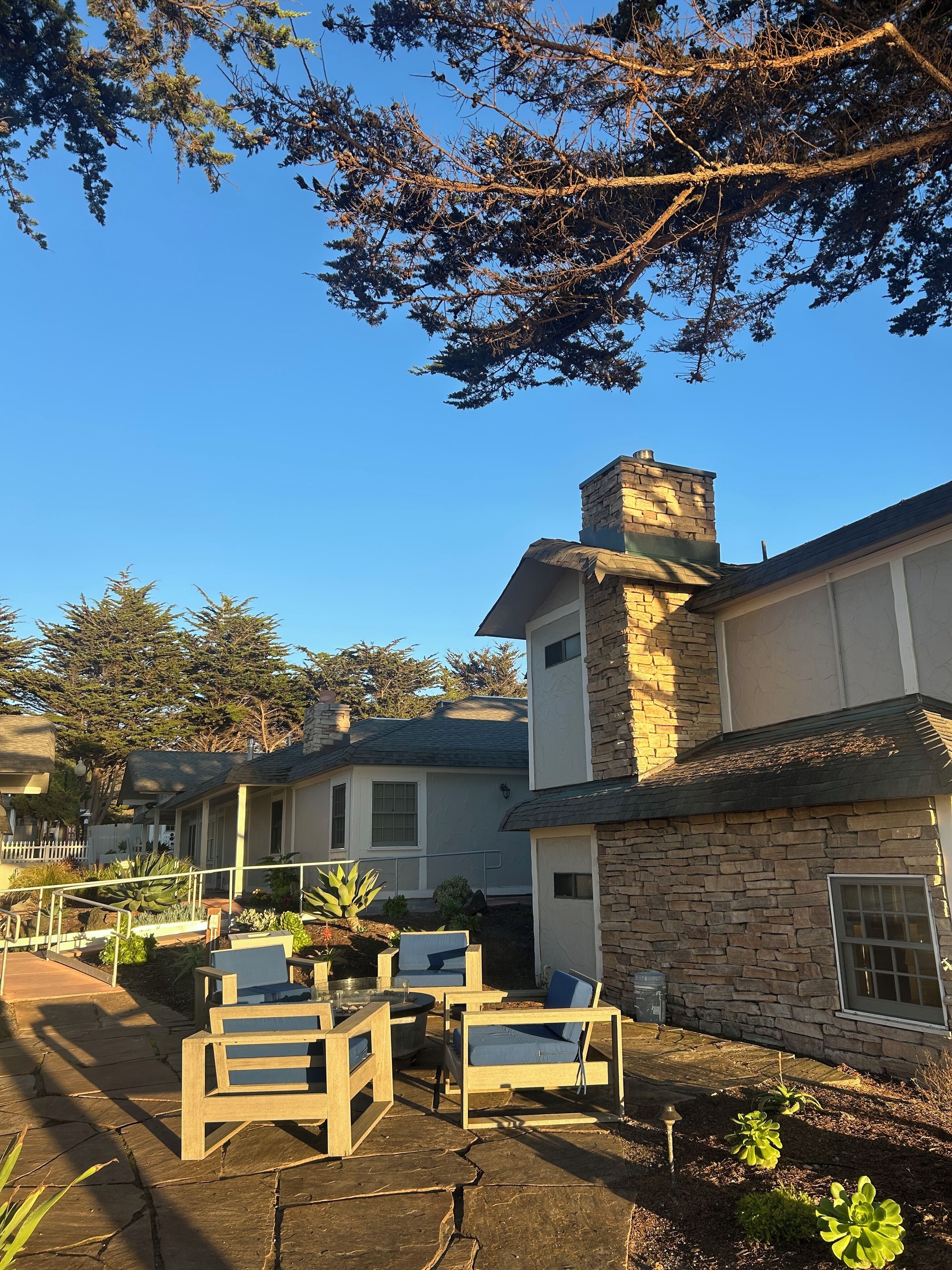 Um imóvel residencial com chaminé de pedra, varanda coberta com mobiliário de madeira e vista para casas vizinhas.