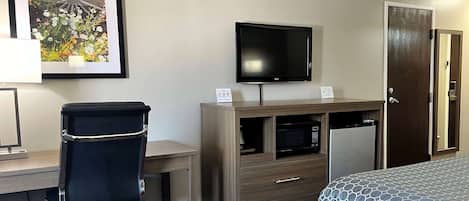 Quarto de hotel com escrivaninha, cadeira, televisão, minibar e cama com colcha estampada.