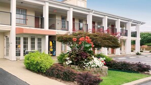 Um edifício de dois andares com varanda frontal coberta, um pequeno jardim com diversas plantas e flores, e uma placa que diz “OFFICE” no andar térreo.