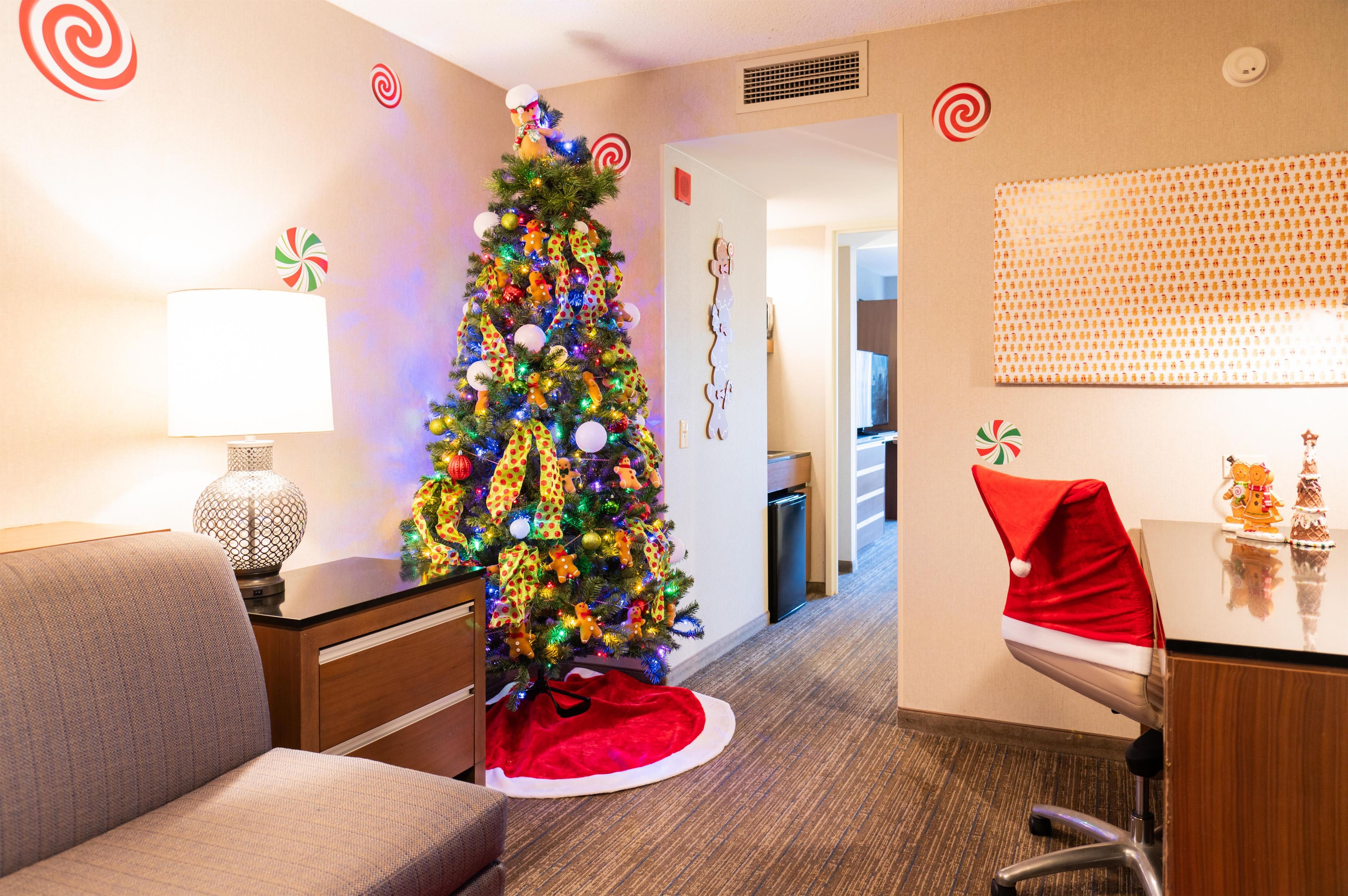 Quarto de hotel com árvore de Natal, uma escrivaninha com abajur, uma cadeira e um espaço de jantar com toalha vermelha.