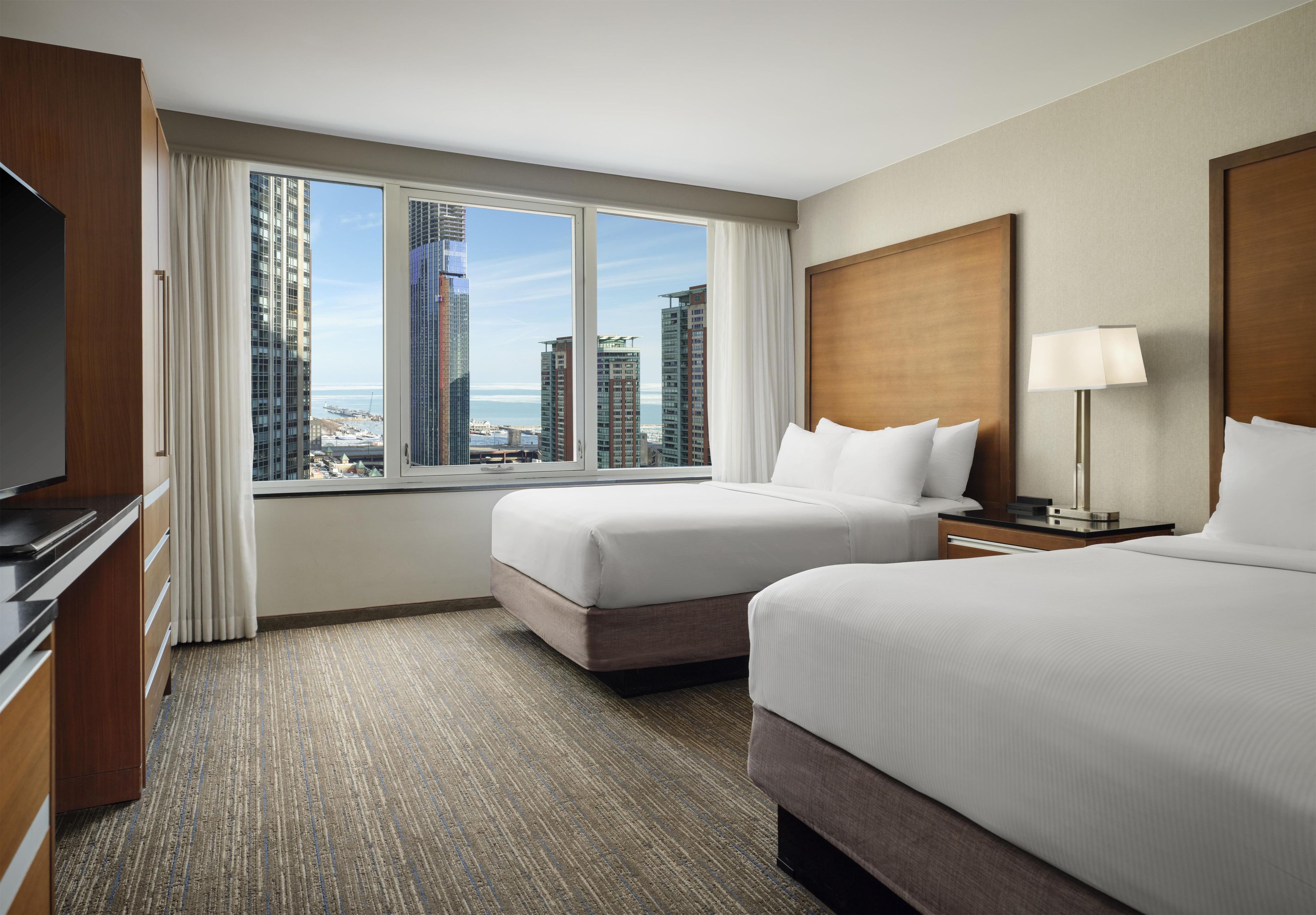 Quarto de hotel com duas camas, televisão, escrivaninha, luminária e vista para edifícios altos.