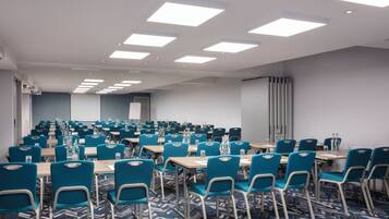 Sala de conferências com fileiras de mesas e cadeiras, equipada com garrafas de água e papéis.
