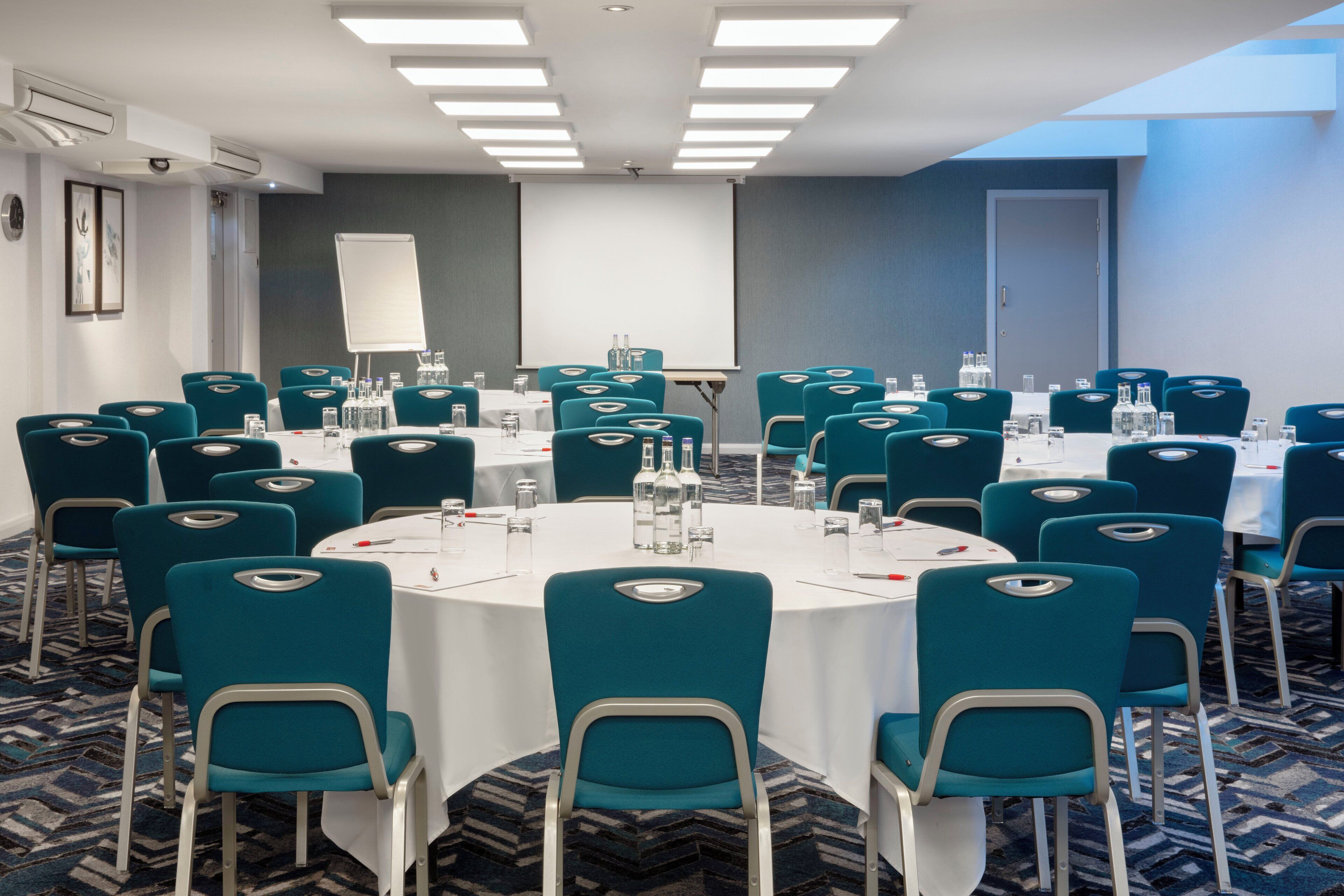 Sala de conferência com cadeiras cor de verde-água e mesas brancas, equipada com garrafas de água e copos, e um quadro branco.