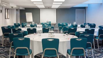 Sala de conferência com cadeiras cor de verde-água e mesas brancas, equipada com garrafas de água e copos, e um quadro branco.