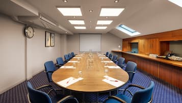 Uma sala de conferências com uma mesa comprida, cadeiras azuis, um relógio e uma tela de projetor.