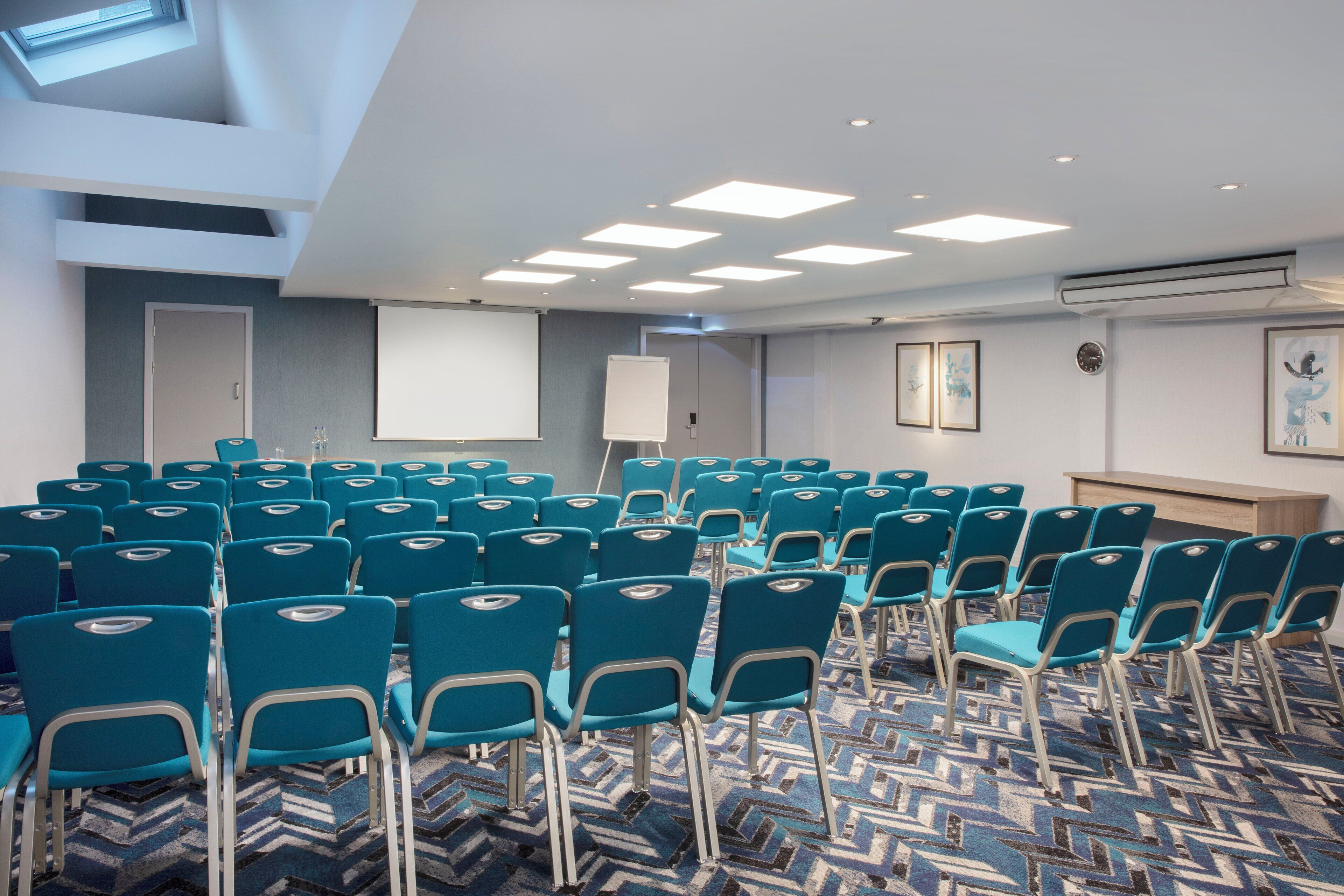 Uma sala de conferências com fileiras de cadeiras azul-esverdeadas, uma lousa branca e uma tela de projetor.