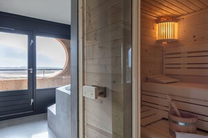 Uma sala de sauna com bancos de madeira, uma porta de vidro que dá para o exterior e um balde de madeira com uma concha.