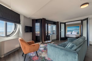 Quarto de hotel moderno com uma janela grande, sofá, poltrona, mesa pequena e cama com vista para a cidade.