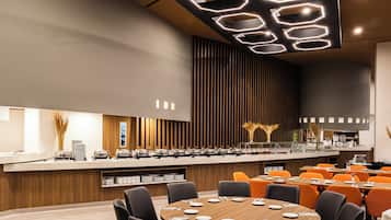 Uma área de refeições moderna com mesas redondas, cadeiras laranja, um balcão de buffet com diversos pratos e um lustre contemporâneo no teto.