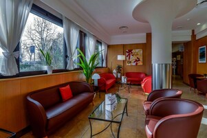 Um saguão de hotel com um sofá vermelho, cadeiras vermelhas, uma mesa de centro de vidro e uma grande janela com cortinas.