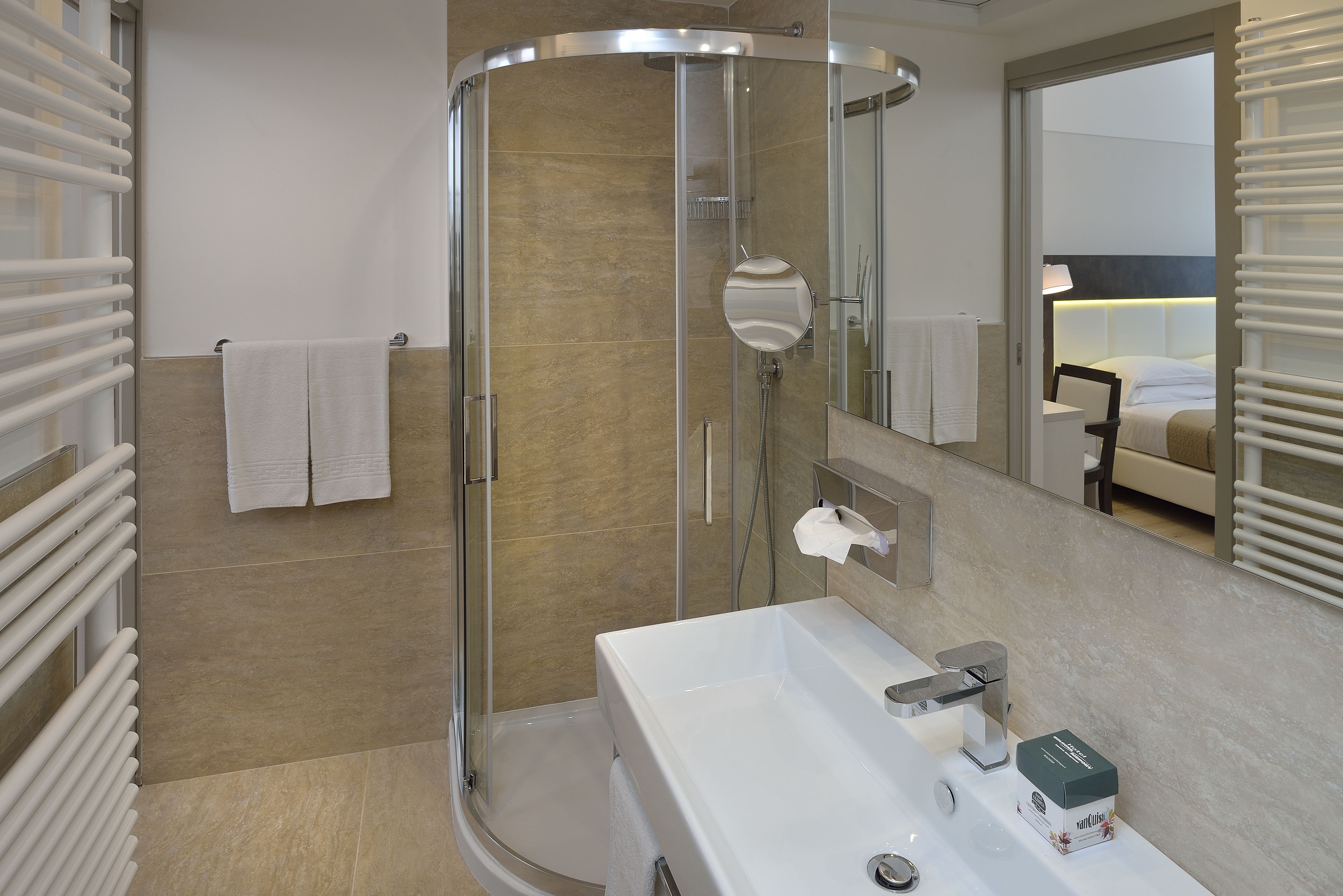 Um banheiro moderno com chuveiro de vidro, pia branca e espelho refletindo um quarto.