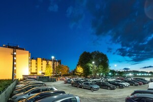 Um estacionamento com vários carros, um edifício com janelas iluminadas e árvores ao fundo.