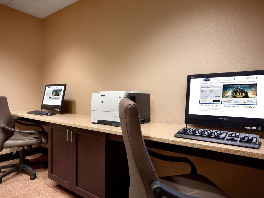 Um espaço de trabalho em hotel, com uma mesa, dois computadores, uma impressora e cadeiras de escritório.