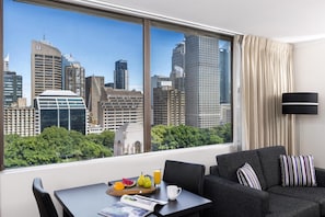 Quarto de hotel com uma grande janela que oferece vista para o horizonte da cidade, uma área de refeições com mesa posta para dois e um sofá de couro preto com almofadas listradas.