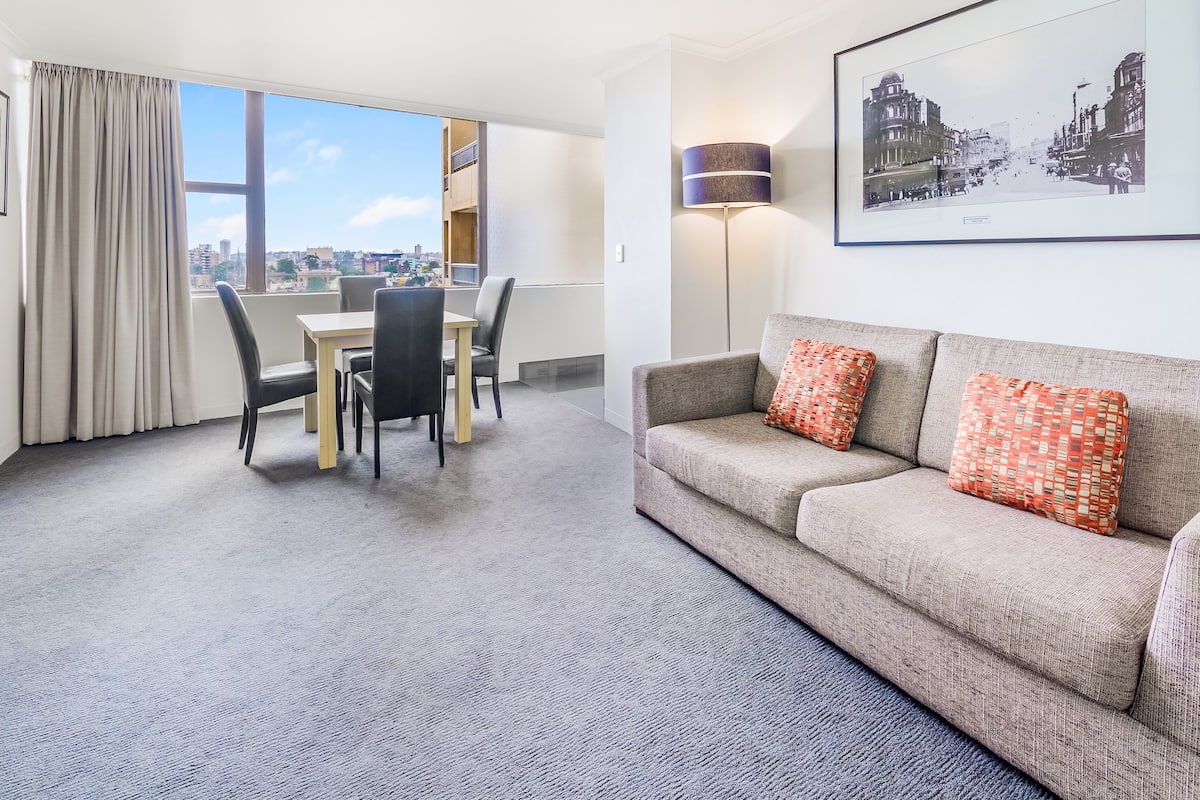 Quarto de hotel com área de refeições, sofá com almofadas estampadas, abajur e uma pintura emoldurada com vista da cidade.
