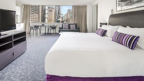 Quarto de hotel com uma cama grande, televisão, escrivaninha e vista para edifícios altos.