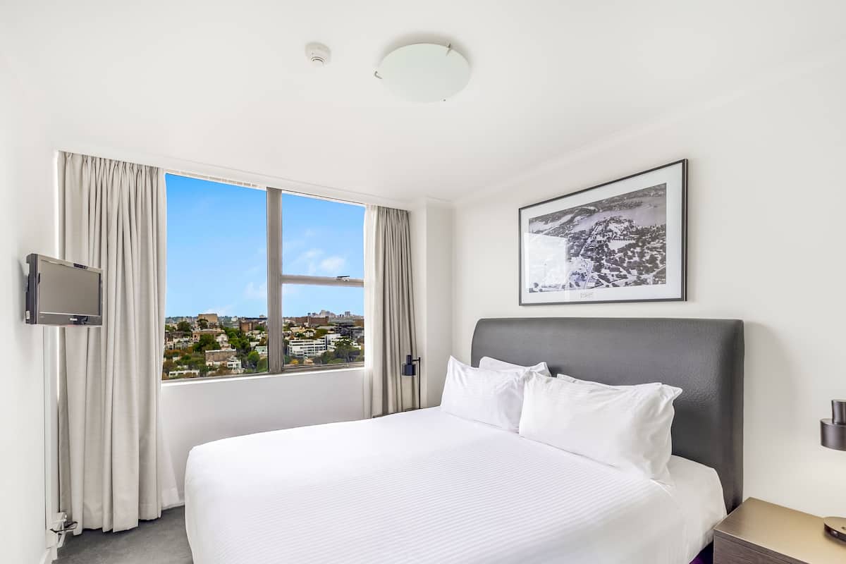 Quarto de hotel com cama grande, televisão de tela plana, mesa de cabeceira, escrivaninha e janela com cortinas.