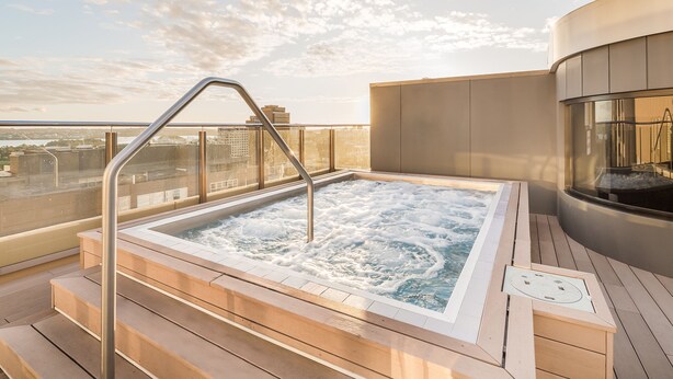 Uma banheira de hidromassagem no terraço, com escada de metal, degraus de madeira e guarda-corpo de vidro.