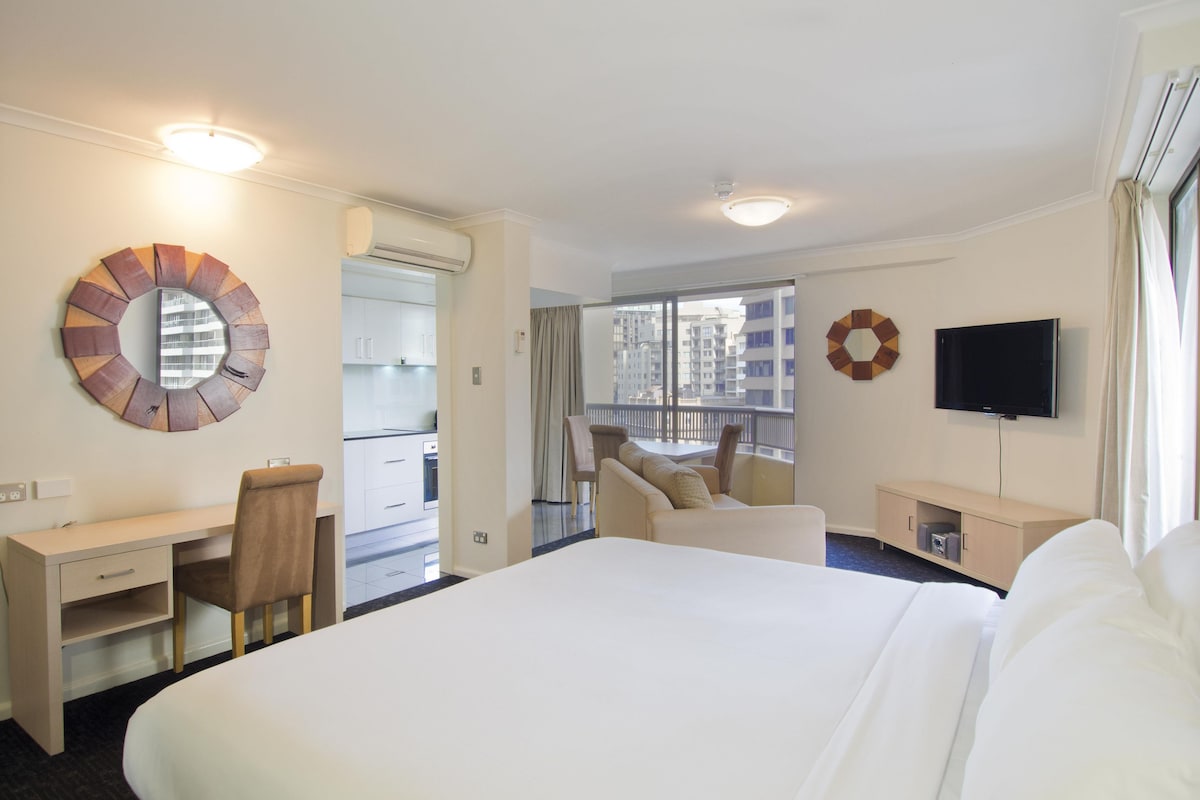 Quarto de hotel com uma cama grande, escrivaninha, cadeira, televisão e vista para a cidade pela janela.