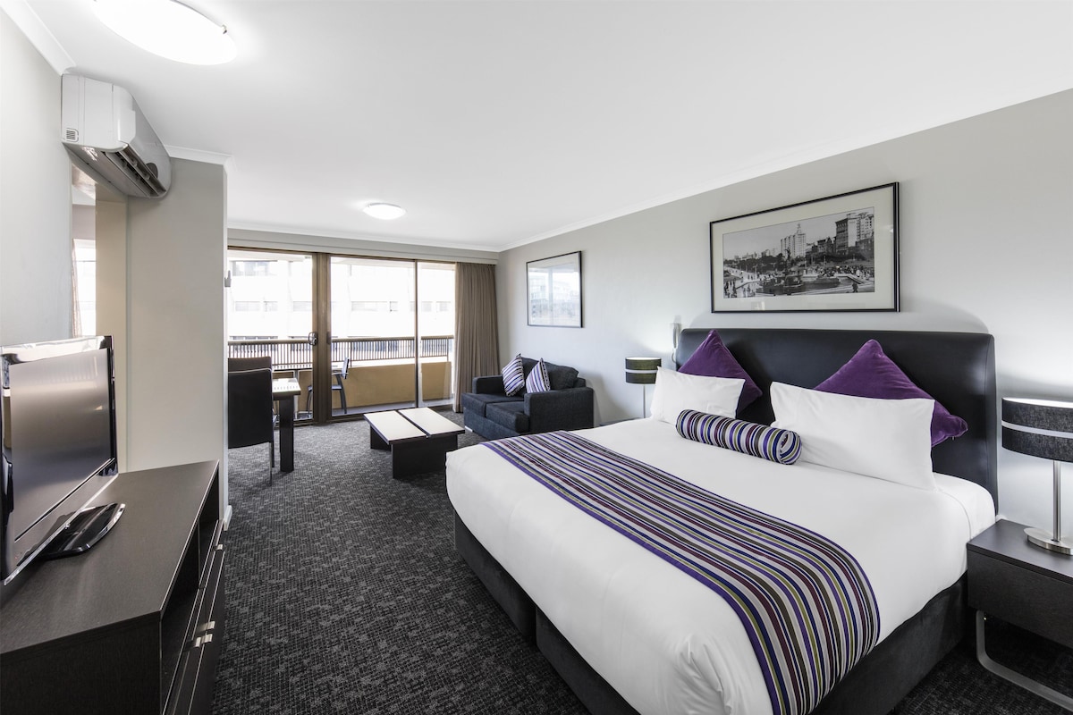 Quarto de hotel com uma cama grande, uma escrivaninha, uma cadeira, televisão, varanda com vista e um quadro na parede.