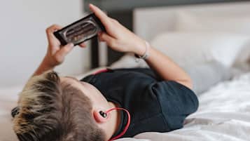 Uma pessoa deitada na cama com um smartphone, vestindo uma camisa preta e uma pulseira vermelha.