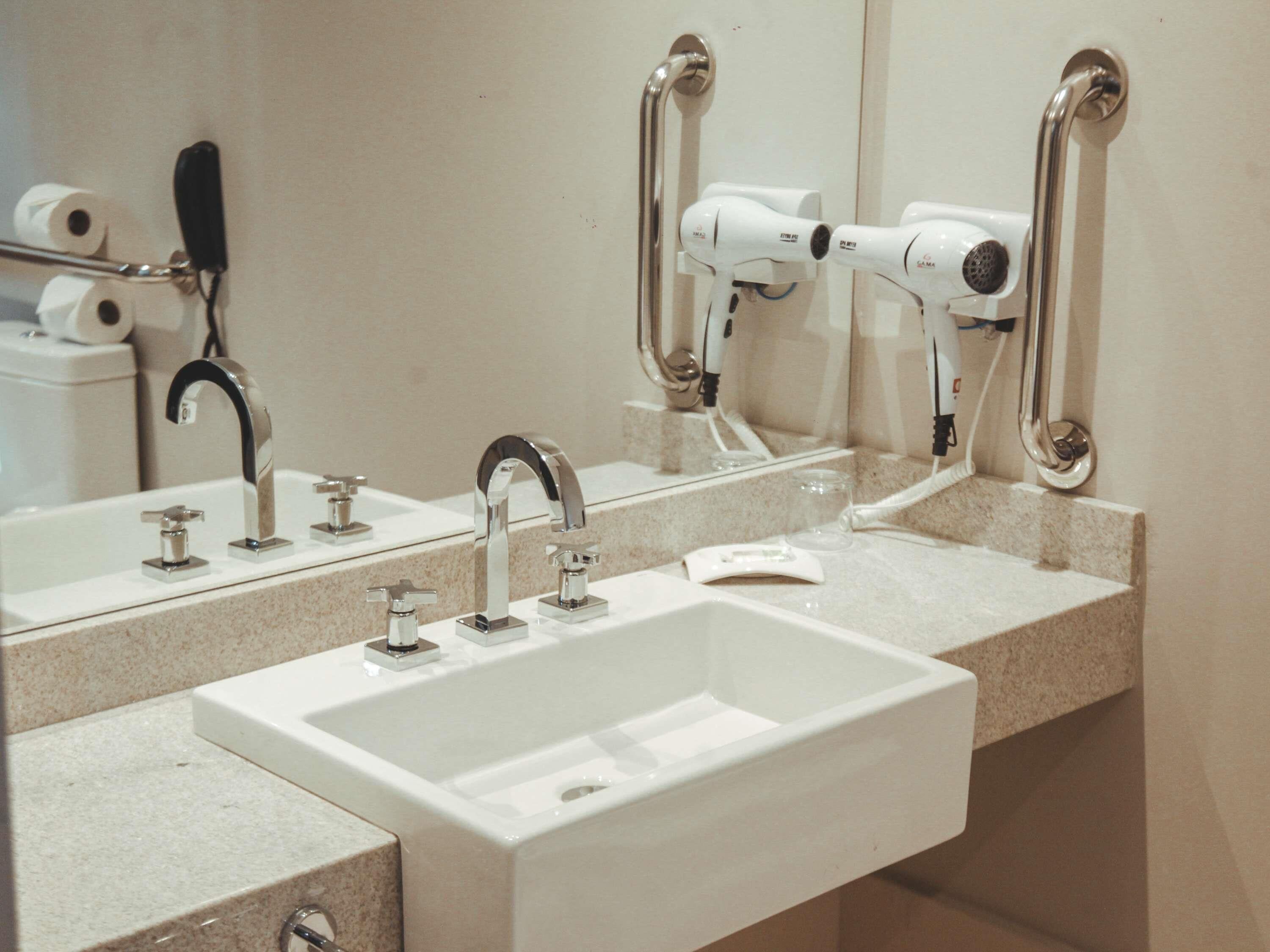 Casa de banho com lavatório branco, torneiras múltiplas, espelho e secador de cabelo fixado na parede.