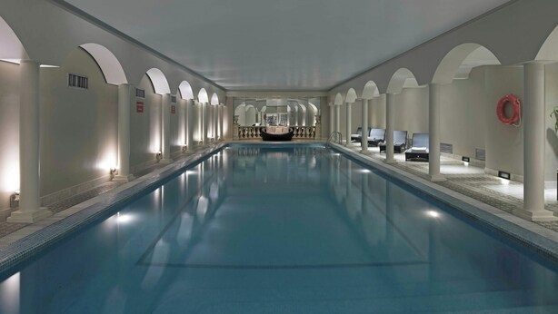 Uma piscina interior comprida e estreita, com uma área de estar central e espreguiçadeiras ao longo das laterais.