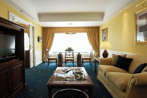Quarto de hotel com mesa de jantar, cadeiras, sofá, televisão e uma janela grande com cortinas.