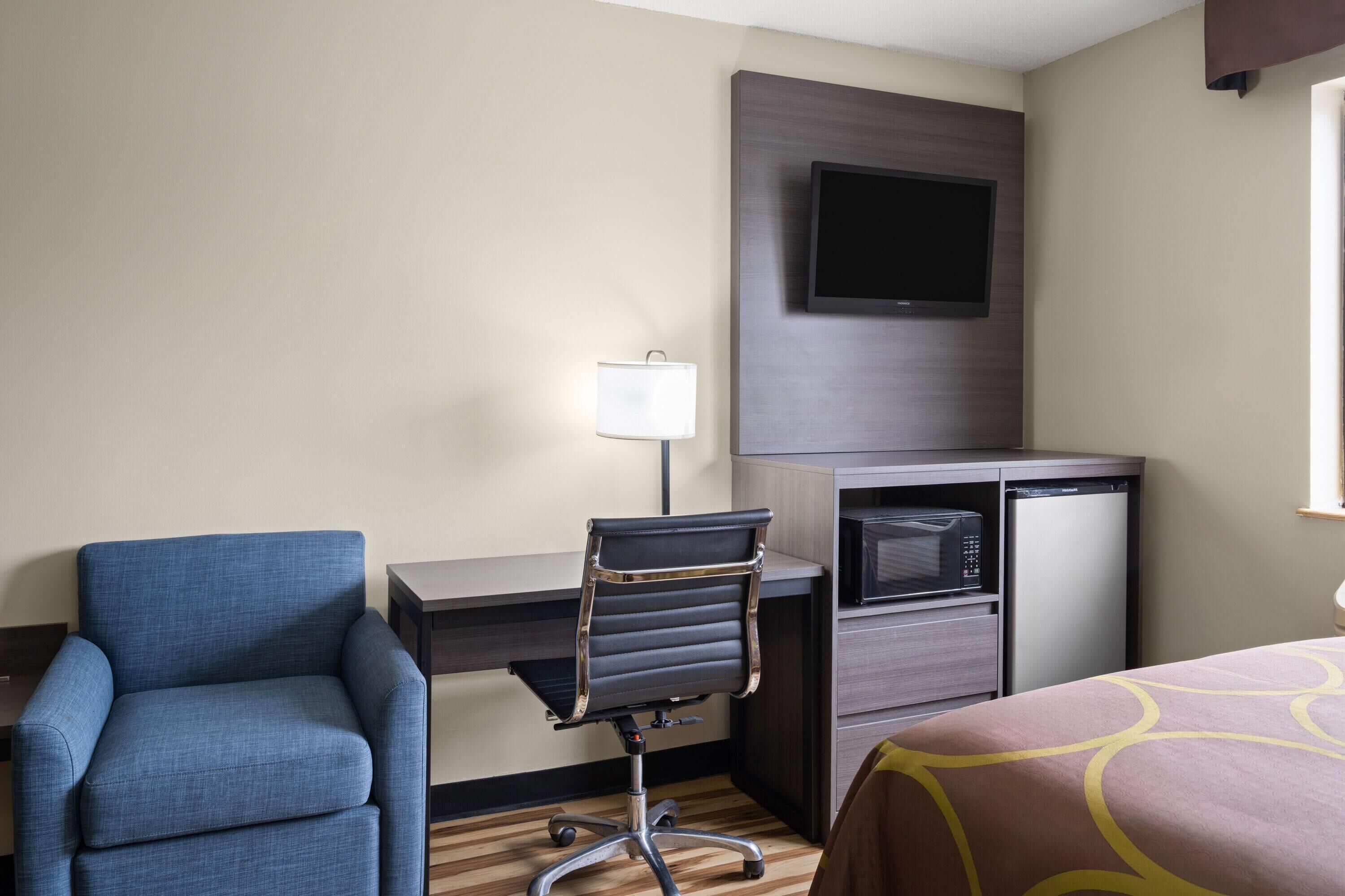 Quarto de hotel com escrivaninha, poltrona azul, televisão na parede, micro-ondas e frigorífico.