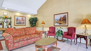 Um hall com um sofá estampado em vermelho, duas poltronas vermelhas, uma mesa de centro de madeira e duas luminárias de chão. Há quadros nas paredes e um vaso com planta.