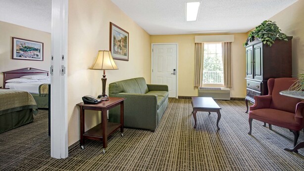 Quarto de hotel com uma cama, um sofá verde, uma mesinha, uma poltrona vermelha e uma televisão.