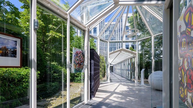 Uma passarela envidraçada com design moderno, rodeada por vegetação e com uma instalação artística central.